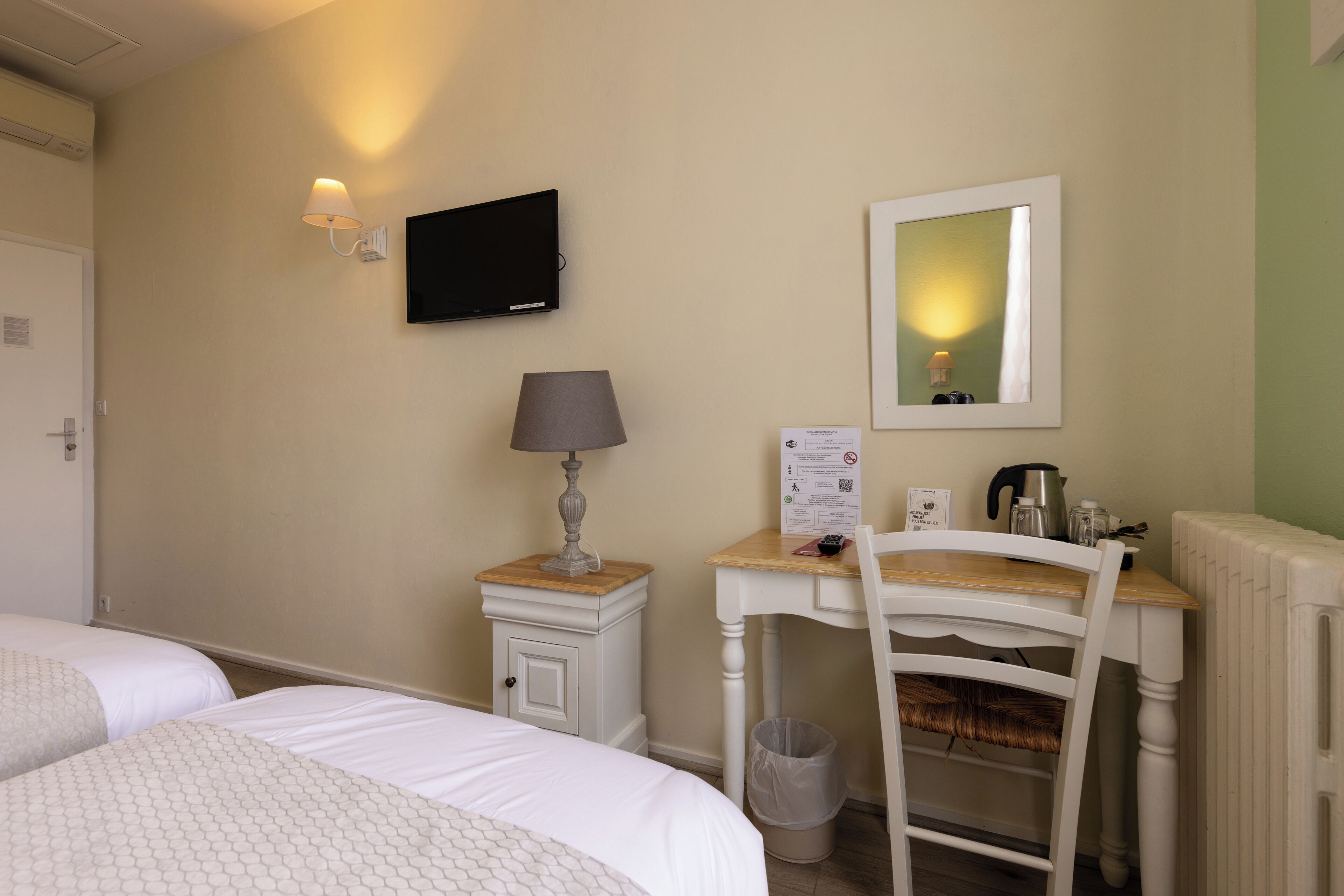 Quarto de hotel com duas camas, uma escrivaninha com cadeira, uma luminária, uma televisão, um espelho e um radiador.