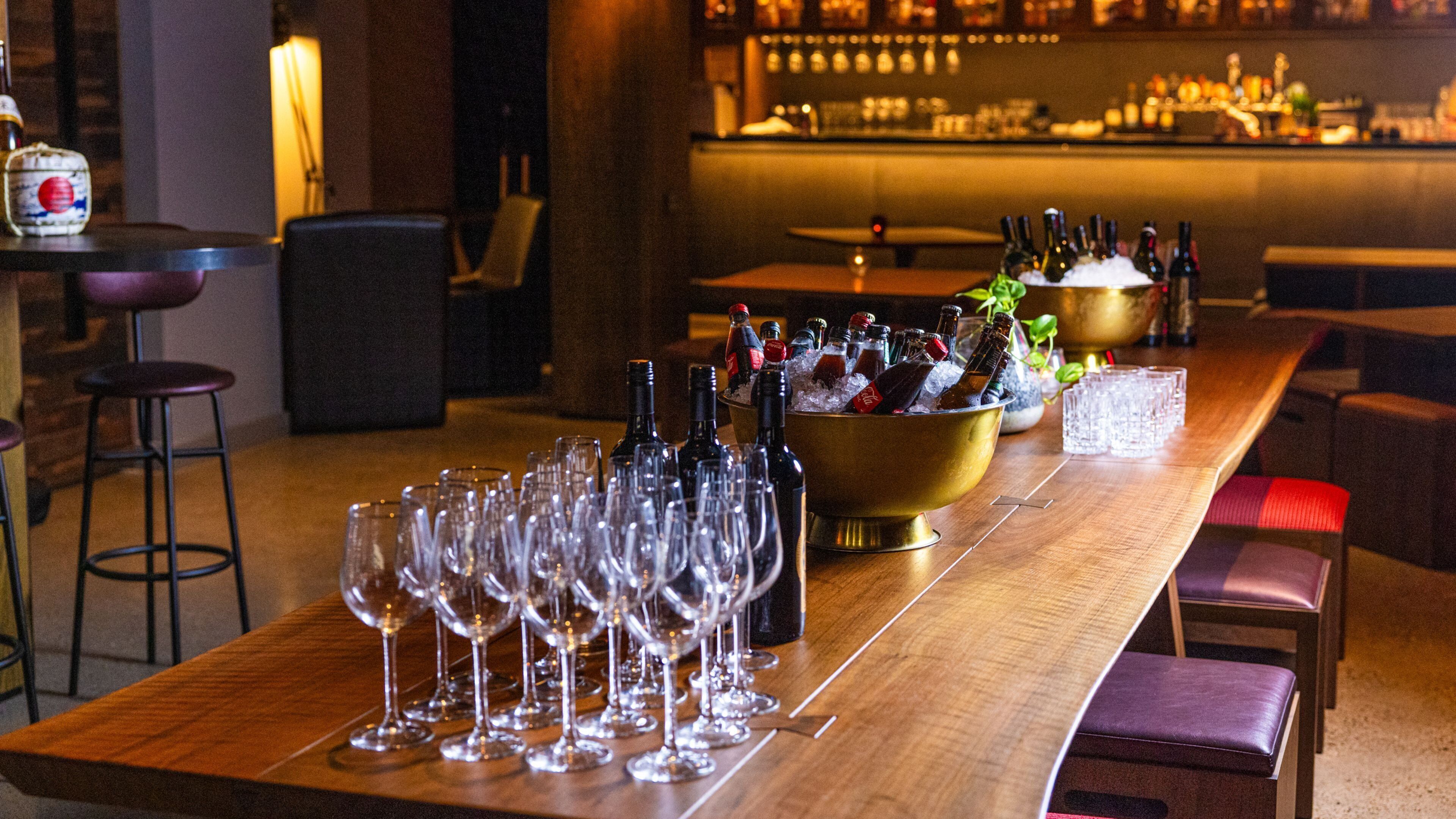 Uma área de bar com um longo balcão de madeira, vários copos de vinho, garrafas de vinho e um balde dourado cheio de gelo e garrafas.