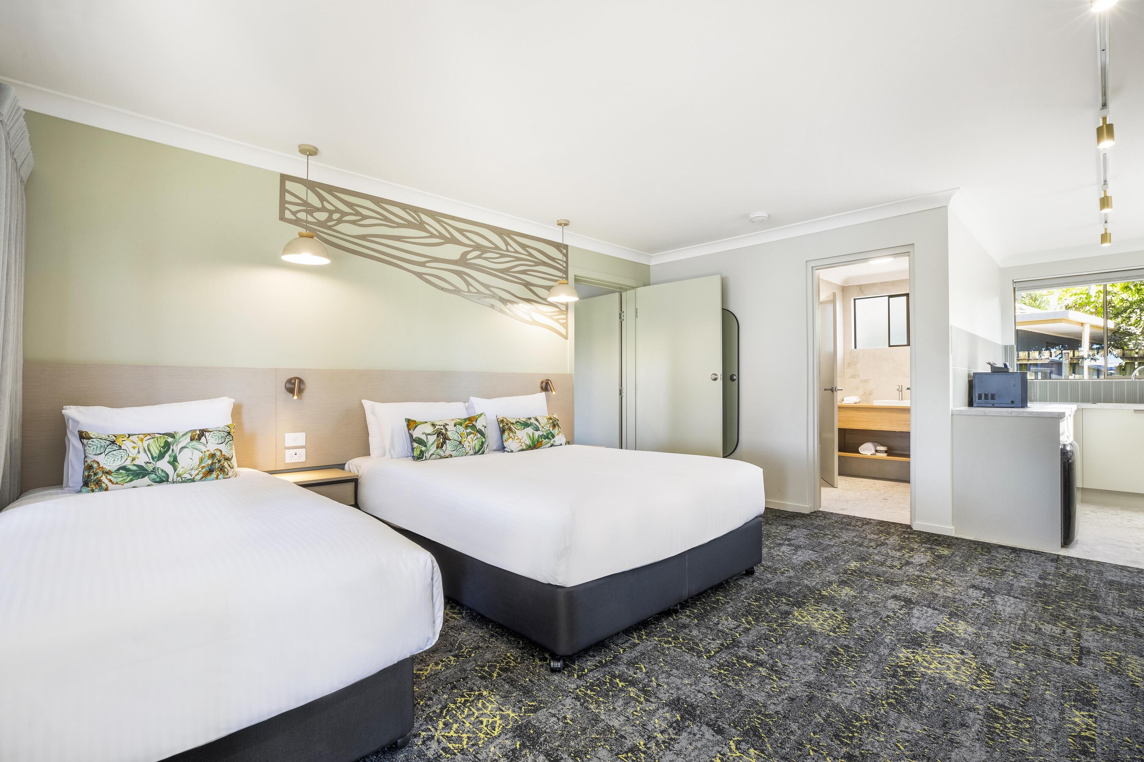 Quarto de hotel com duas camas, um tapete escuro estampado, um espelho grande e vista para o banheiro.