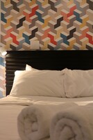 Uma cama bem arrumada com lençóis e almofadas brancas, cabeceira e papel de parede estampado ao fundo.