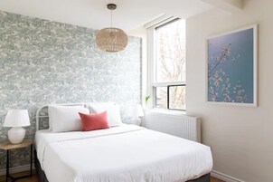 Quarto com cama, mesa de cabeceira, luminária, janela com vista para árvores e parede com papel de parede floral.