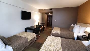 Quarto de hotel com duas camas de solteiro, uma mesa com cadeira, televisão fixada na parede e uma luminária.