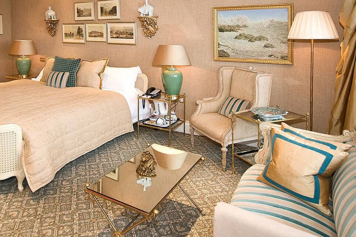 Quarto de hotel com uma cama grande, um espaço de estar com sofá e poltrona, uma mesinha e um banheiro visível através de uma porta aberta.