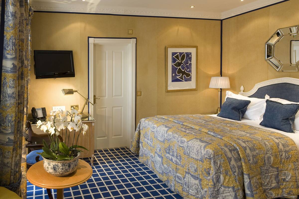 Quarto de hotel com cama, escrivaninha com luminária, planta em vaso, televisão e porta.