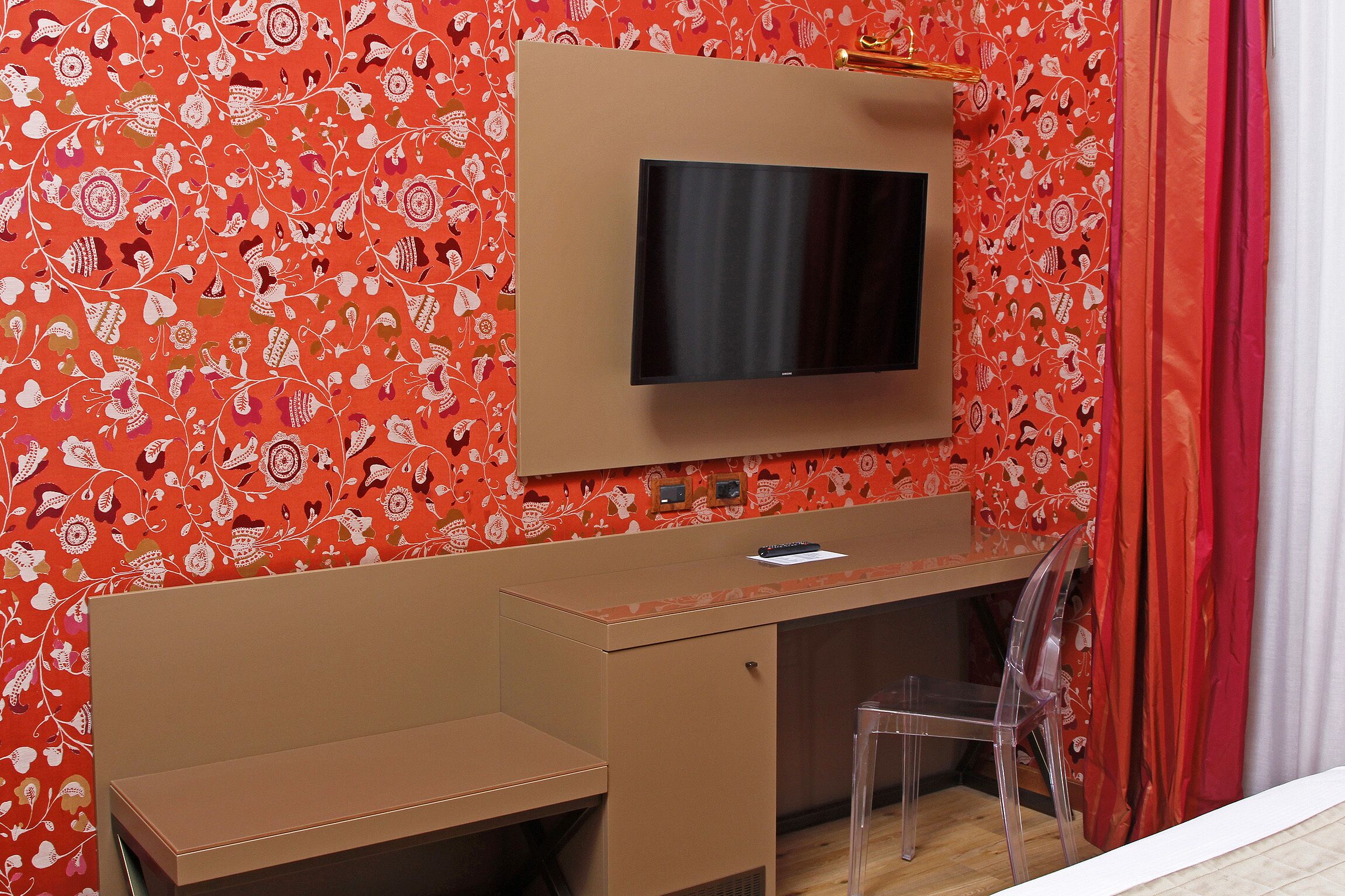 Quarto com televisão na parede, uma escrivaninha e uma cadeira, e papel de parede estampado em vermelho.