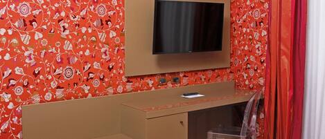 Quarto com televisão na parede, uma escrivaninha e uma cadeira, e papel de parede estampado em vermelho.