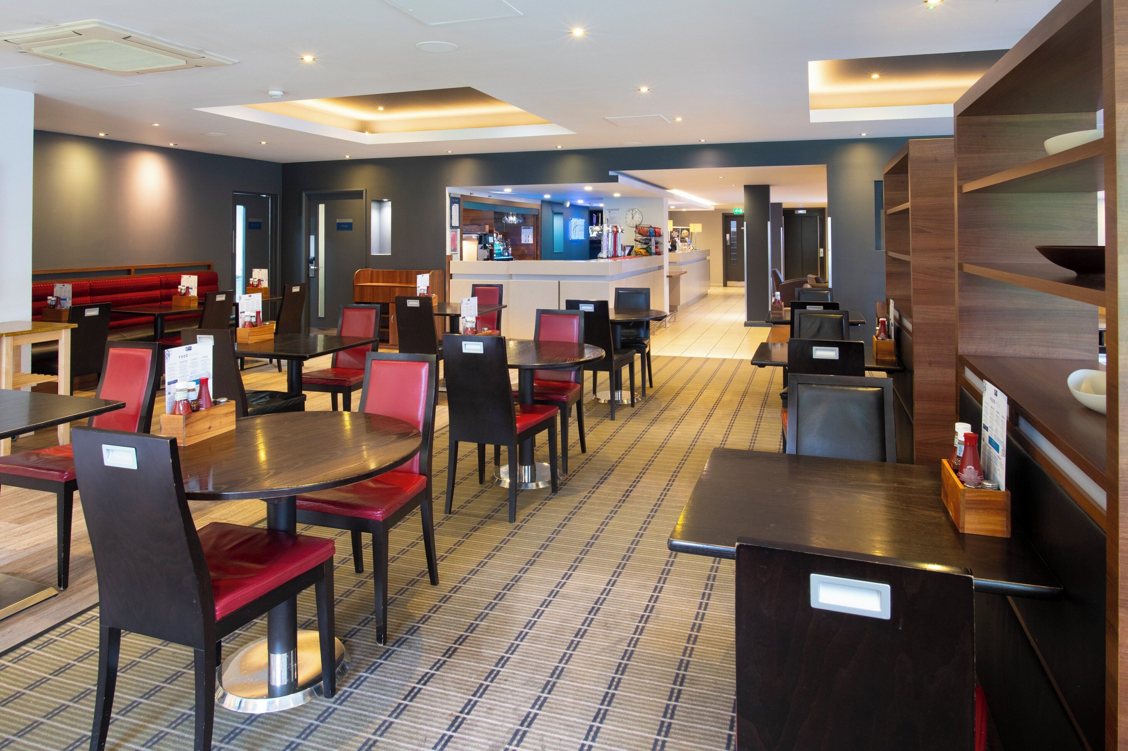 Uma área de refeições de hotel com várias mesas e cadeiras, um bar e uma receção.