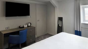 Quarto de hotel com cama, escrivaninha com cadeira, televisão na parede e janela com cortinas.