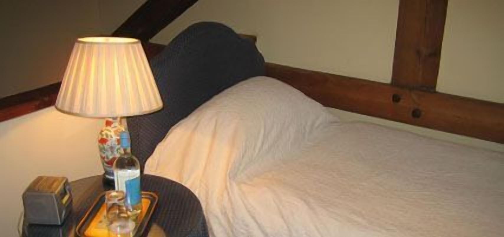 Cama bem arrumada com edredom branco e travesseiro azul, abajur, mesinha com vaso e garrafa, e cabeceira de madeira.