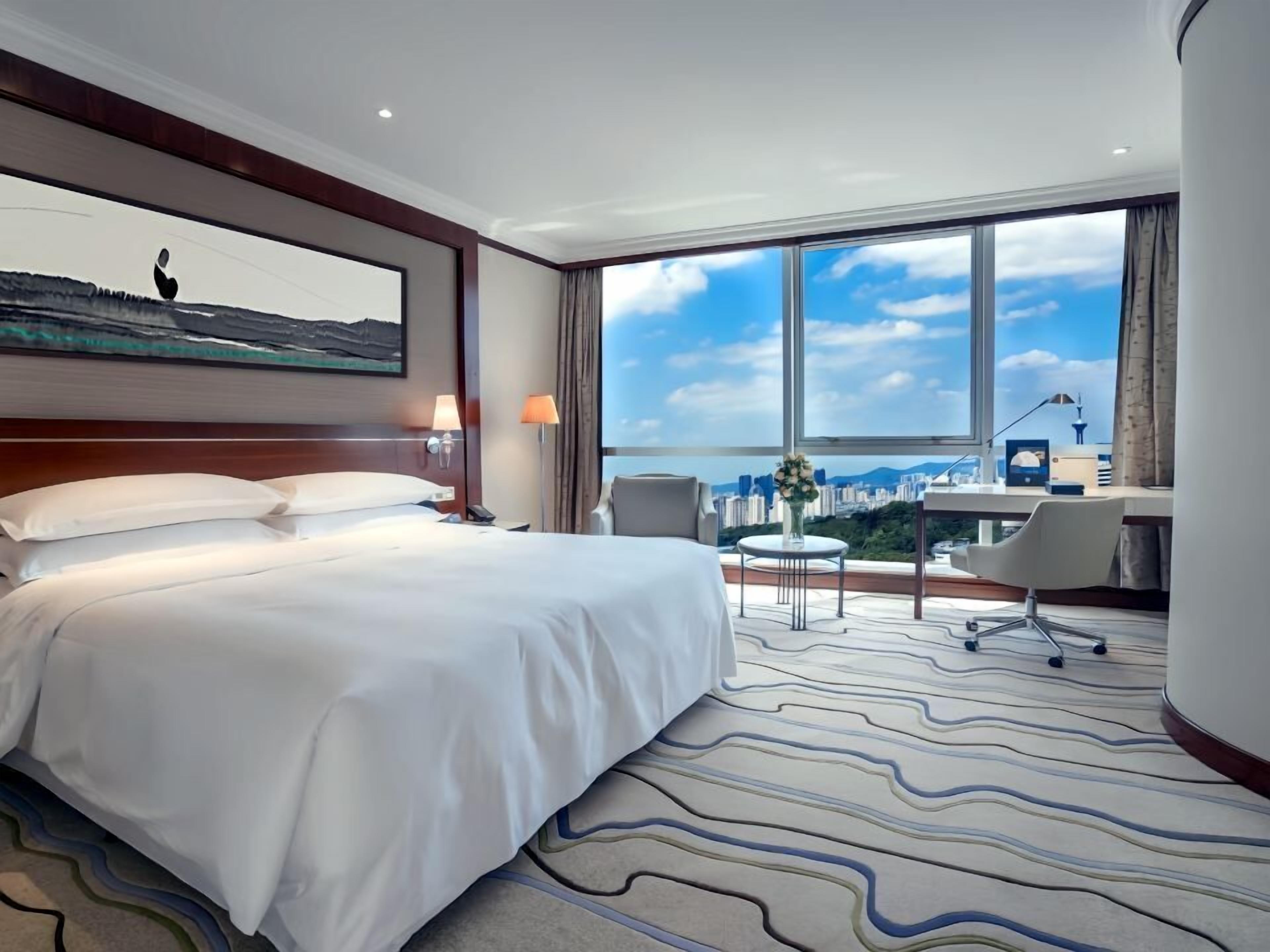 Quarto de hotel com uma cama grande, uma mesa com cadeira, vista da cidade e um quadro na parede.