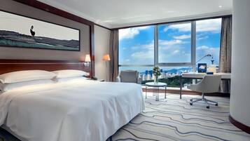 Quarto de hotel com uma cama grande, uma mesa com cadeira, vista da cidade e um quadro na parede.