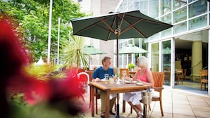 Um pátio externo com mesa e cadeiras de madeira, um guarda-sol e um edifício de vidro ao fundo.