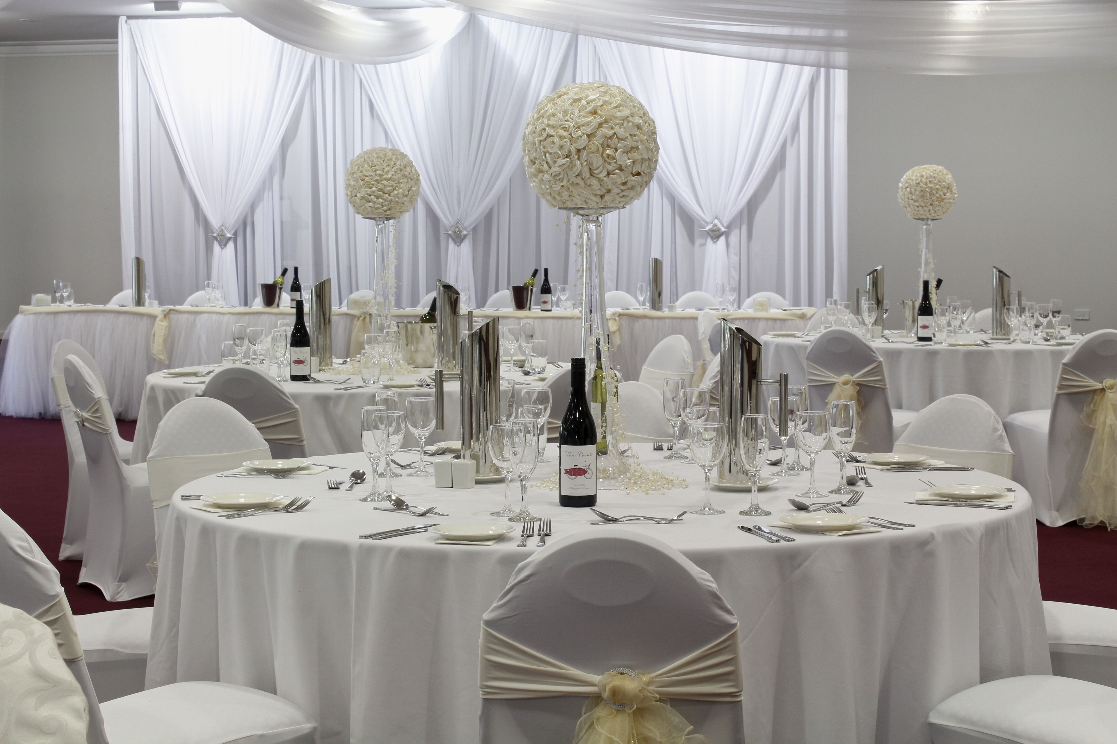 Uma sala de banquetes com mesas redondas preparadas para um evento formal, cada uma com toalhas brancas, talheres e garrafas de vinho.
