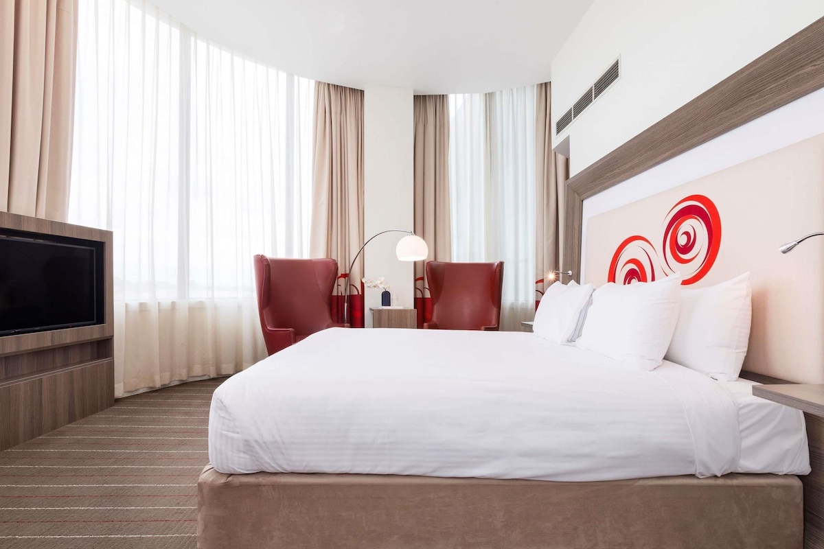 Quarto de hotel com uma cama grande, duas cadeiras vermelhas, uma televisão e uma parede com desenhos circulares vermelhos.