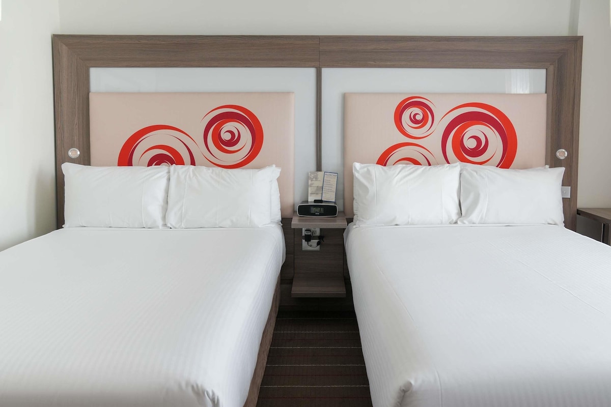 Quarto de hotel com duas camas, uma cabeceira de madeira, uma mesinha de cabeceira com telefone e uma parede com arte abstrata em vermelho e branco.