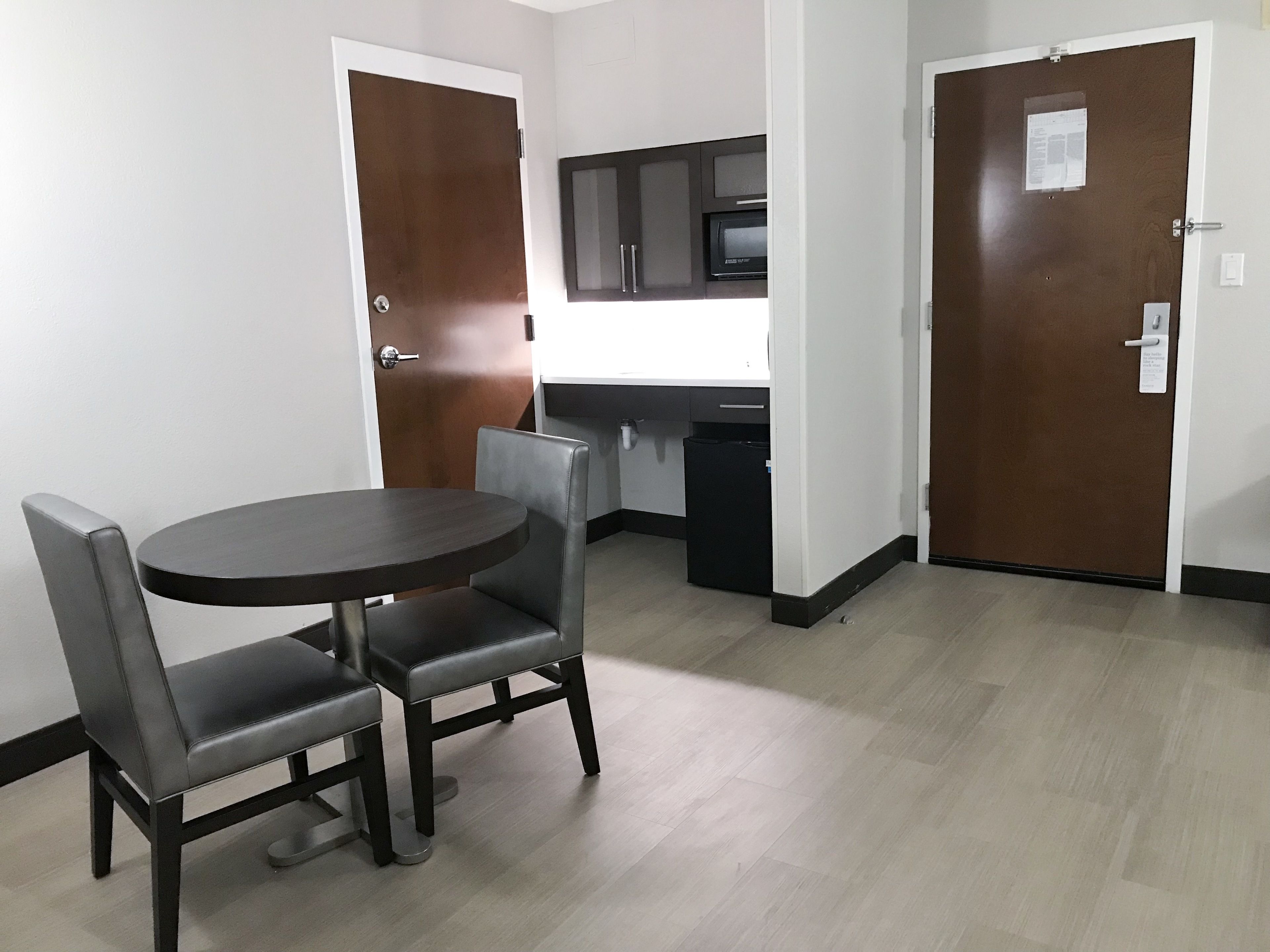 Uma área de refeições com uma mesa redonda e duas cadeiras, uma cozinha com armários e micro-ondas, e duas portas, uma delas com uma placa.