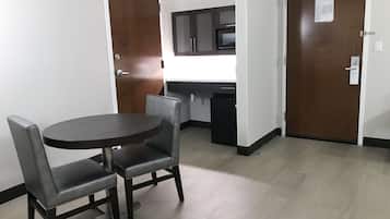 Uma área de refeições com uma mesa redonda e duas cadeiras, uma cozinha com armários e micro-ondas, e duas portas, uma delas com uma placa.