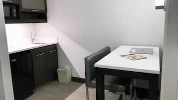 Uma pequena cozinha compacta com micro-ondas, pia e armários pretos. Há uma mesa branca com uma cadeira e um cesto de lixo pequeno.