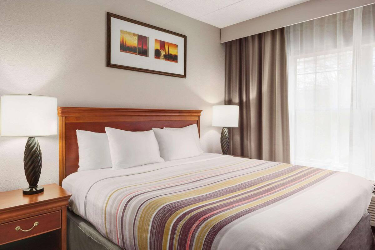 Quarto de hotel com uma cama grande, duas luminárias de cabeceira, um quadro na parede e uma janela com cortinas.