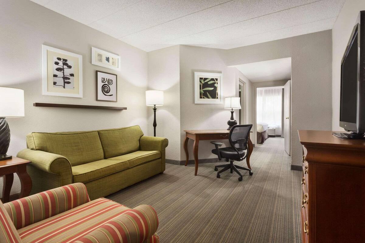 Quarto de hotel com um sofá verde, uma poltrona listrada, uma secretária com cadeira e uma televisão sobre um aparador de madeira.