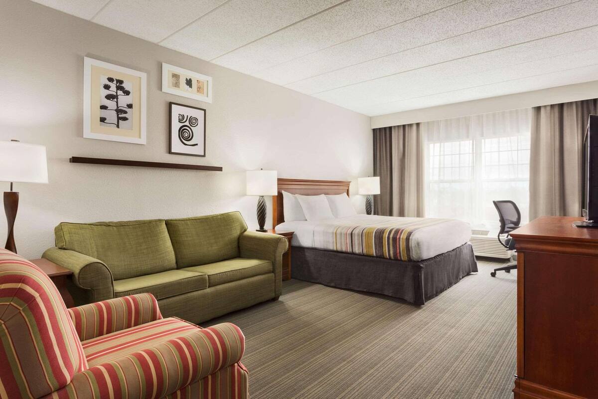 Quarto de hotel com uma cama, um sofá verde, uma poltrona listrada e uma escrivaninha com luminária.