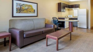 Quarto de hotel compacto com sofá, pequena mesa de jantar, cadeira, cozinha equipada com micro-ondas e frigorífico, e um quadro na parede.