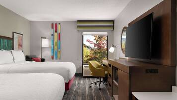 Quarto de hotel com duas camas, uma mesa, uma cadeira, uma televisão e janela com vista para árvores.