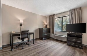 Quarto de hotel com escrivaninha, cadeira, televisão, cômoda e janela com cortinas.