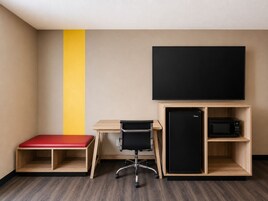Quarto compacto com uma escrivaninha, cadeira de escritório preta, um banco vermelho, televisão preta e refrigerador preto.