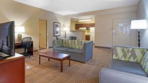 Quarto de hotel com televisão, escrivaninha, sofá, mesa de centro e cozinha compacta ao fundo.