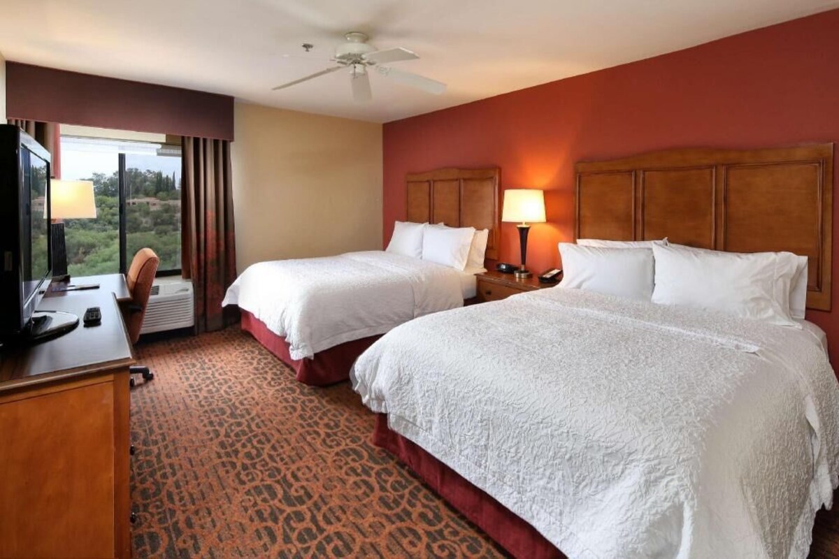 Quarto de hotel com duas camas, uma escrivaninha com computador, uma cadeira, televisão, ventoinha no teto e janela com cortinas.
