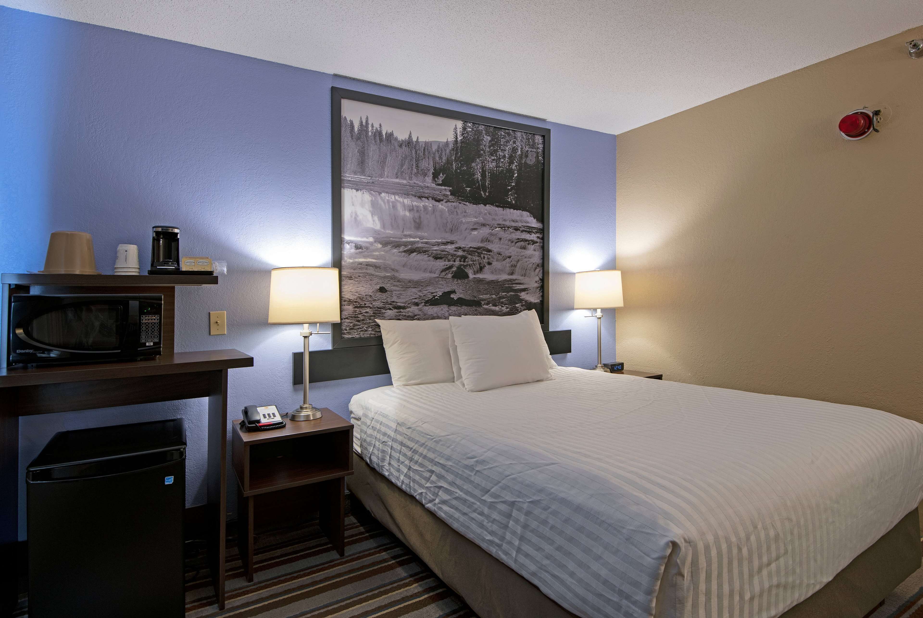 Quarto de hotel com cama, escrivaninha com micro-ondas, cafeteira, luminária e quadro de uma cascata na parede.