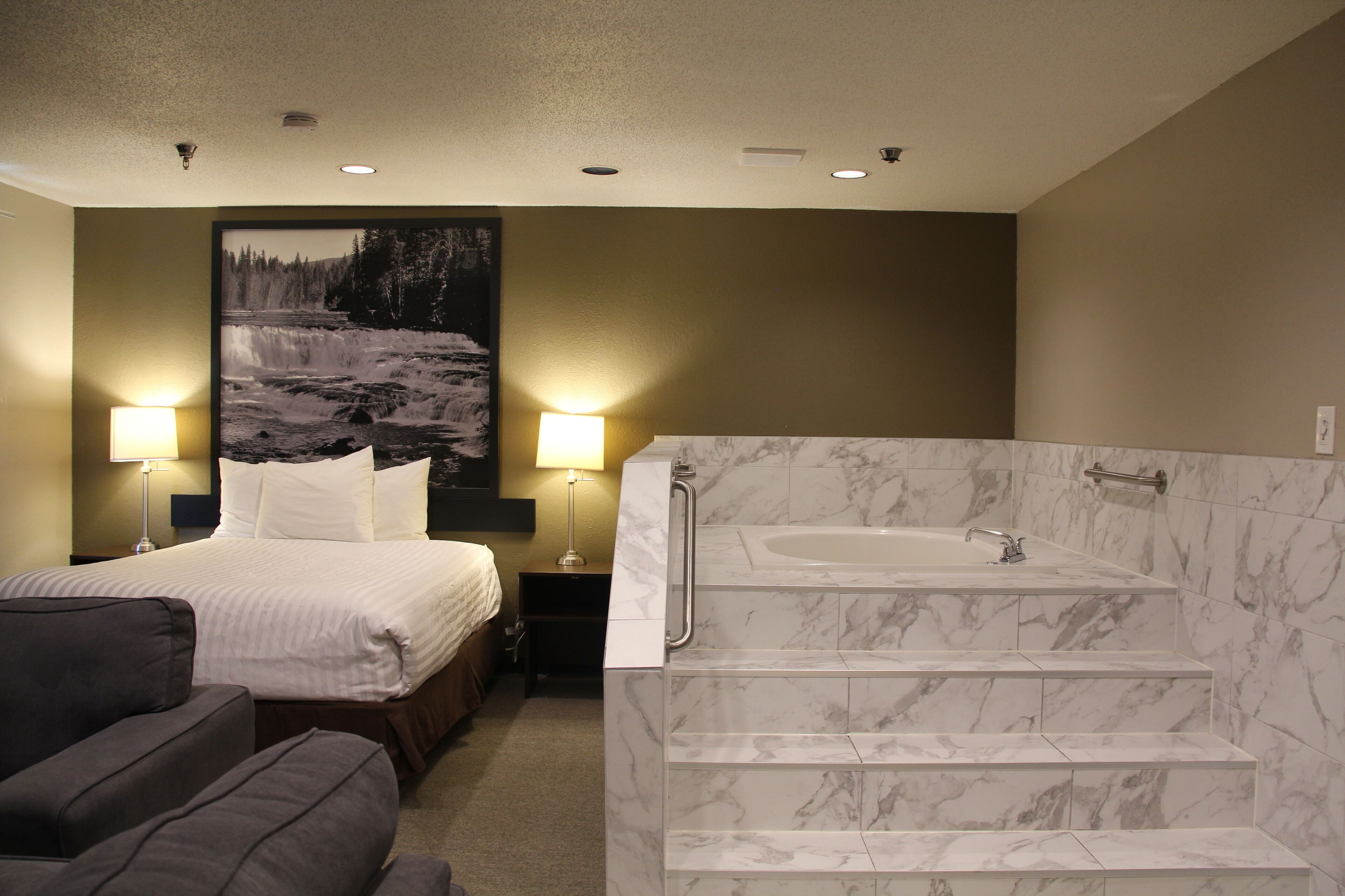 Quarto de hotel com cama, uma grande pintura de uma cascata, banheiro com banheira de mármore e degraus, e duas luminárias de cabeceira.