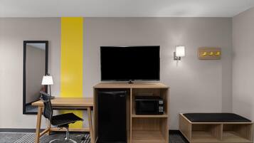 Quarto de hotel com escrivaninha, cadeira, TV, frigorífico, micro-ondas e um espaço de estar pequeno.