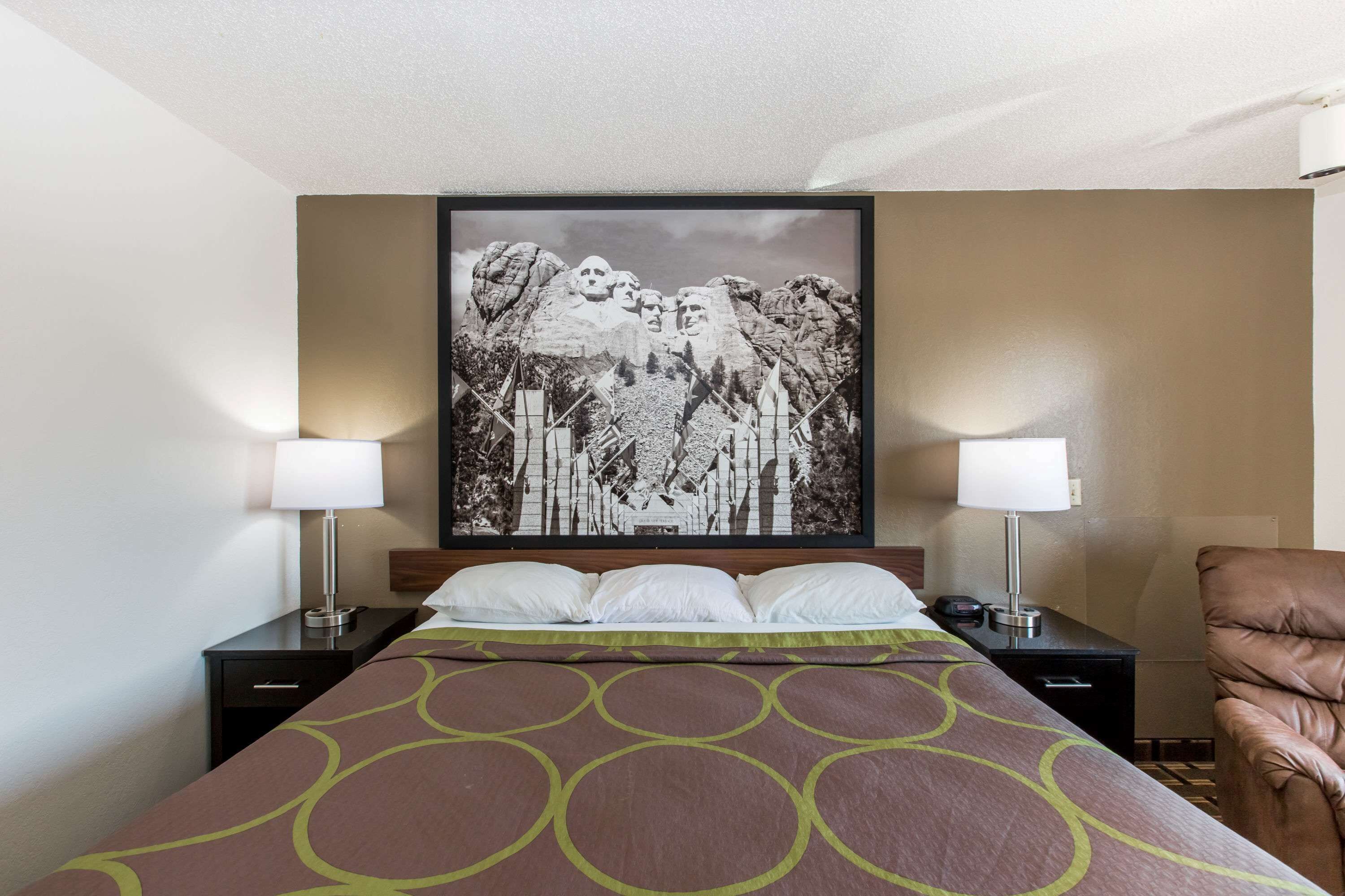 Quarto de hotel com uma cama, duas luminárias laterais, um edredom marrom estampado, um quadro com uma paisagem de montanhas e uma poltrona de couro marrom.