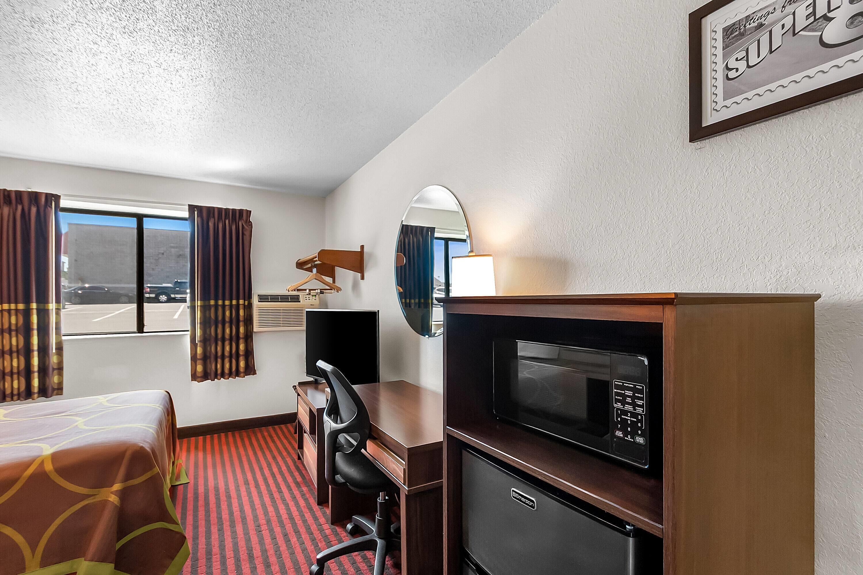 Quarto de hotel com cama, escrivaninha, cadeira, micro-ondas, frigorífico e uma janela grande com cortinas.