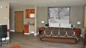Quarto de hotel com cama, escrivaninha, cadeira, pequeno espaço de cozinha e uma janela grande com vista para árvores.