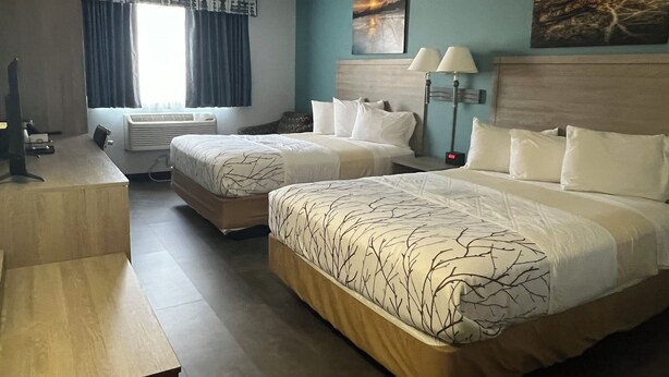 Quarto de hotel com duas camas, uma escrivaninha, uma cadeira, ar condicionado, uma luminária e uma janela com cortinas.