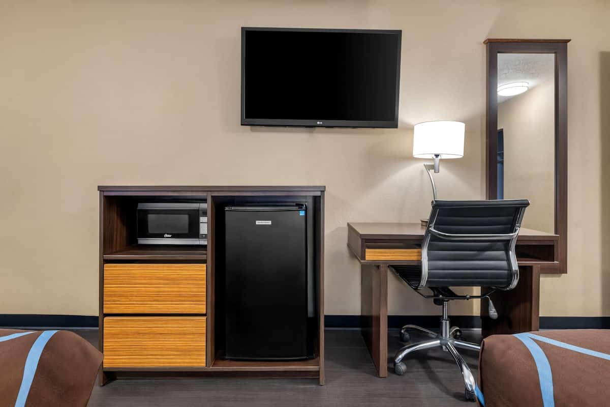 Quarto de hotel com televisão de tela plana na parede, micro-ondas, geladeira, escrivaninha, cadeira e mesa de cabeceira com abajur.