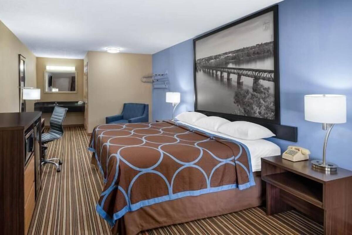 Quarto de hotel com cama, escrivaninha, cadeira, televisão e um quadro na parede.