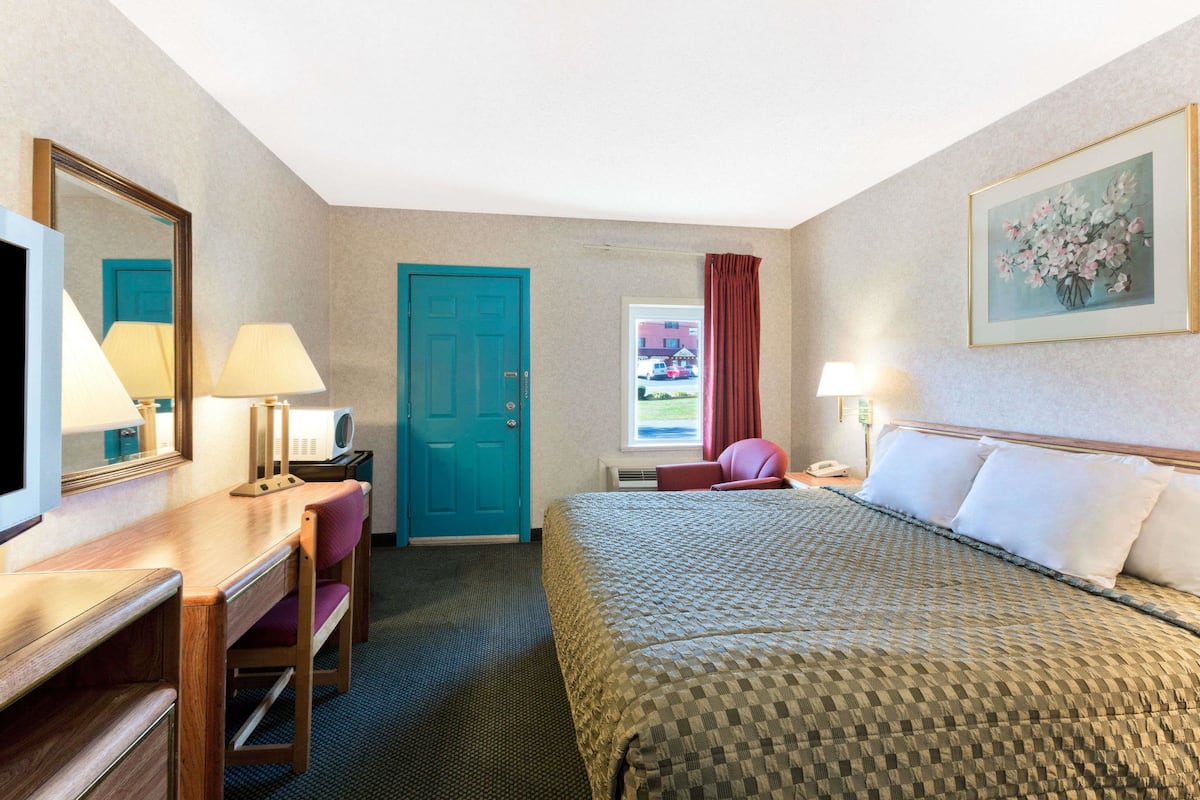Quarto de hotel com cama, escrivaninha com cadeira, televisão, porta e janela com vista para o exterior.