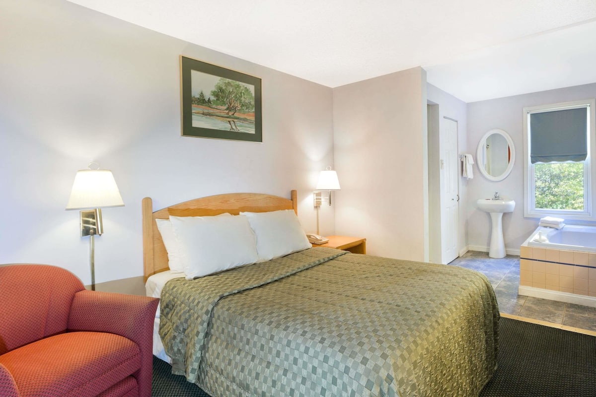 Quarto de hotel com cama, poltrona, mesa de cabeceira com luminária, janela, pia e espelho.
