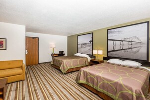 Quarto de hotel com duas camas, um sofá, uma escrivaninha e uma luminária.