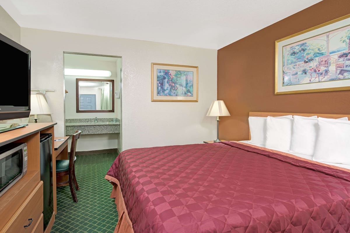 Quarto de hotel com cama, escrivaninha com televisão, cadeira, espelho e um quadro na parede.
