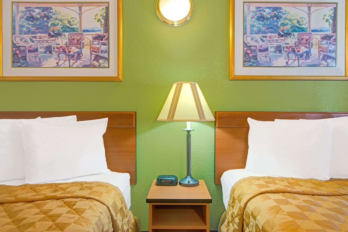 Quarto de hotel com duas camas, uma mesa de cabeceira com abajur, e duas obras de arte emolduradas nas paredes.