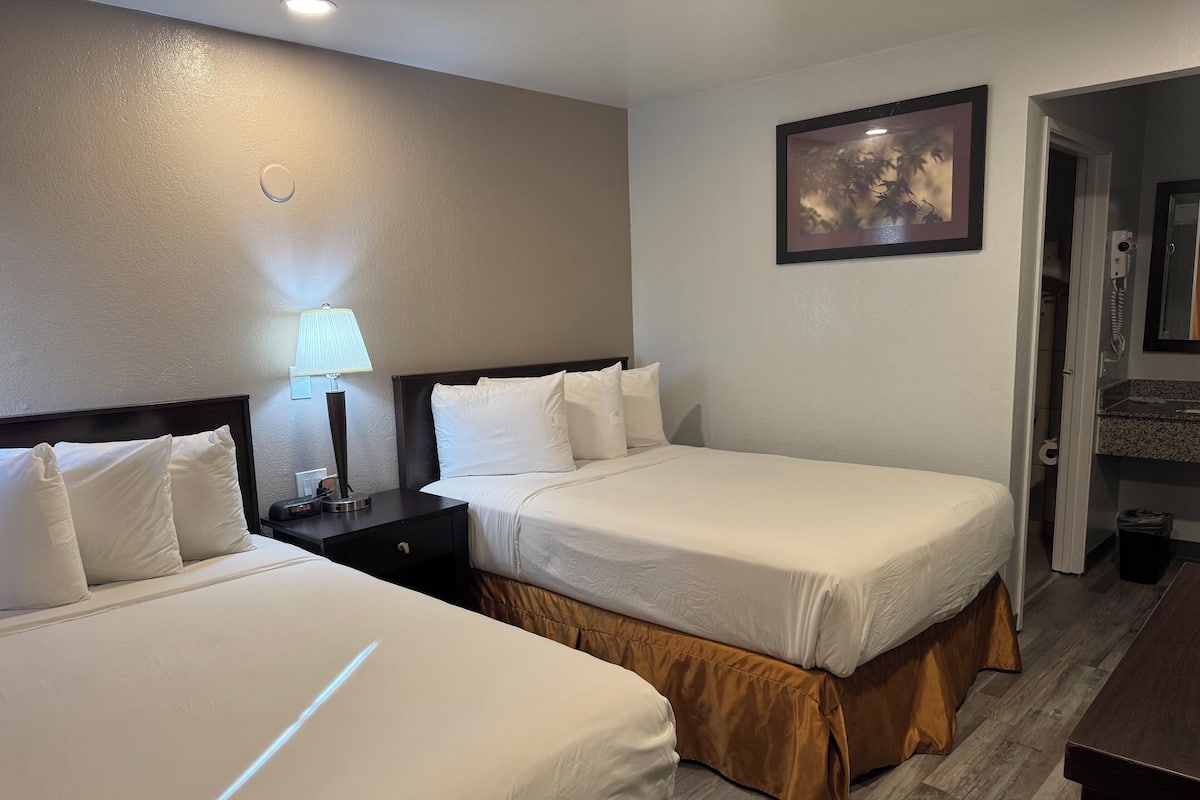 Quarto de hotel com duas camas, uma mesa de cabeceira com abajur, um quadro na parede e uma porta para o banheiro.