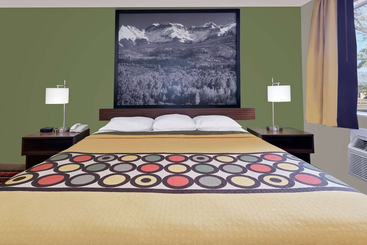 Quarto de hotel com cama, duas mesas de cabeceira, um criado-mudo, um quadro grande com paisagem de montanhas e uma janela com cortinas.