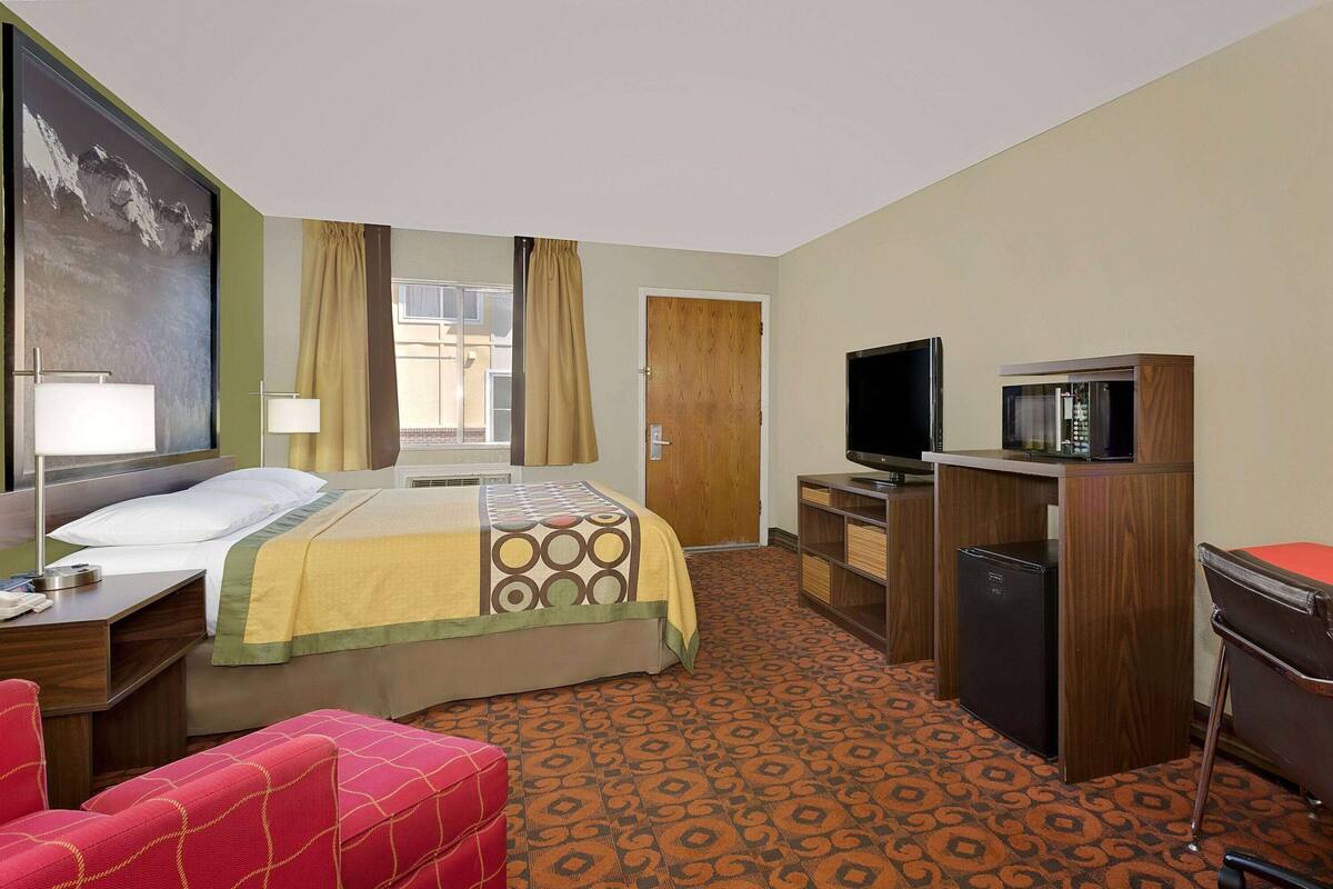 Quarto de hotel com uma cama, uma escrivaninha com televisão e micro-ondas, uma poltrona vermelha e uma janela grande com cortinas.