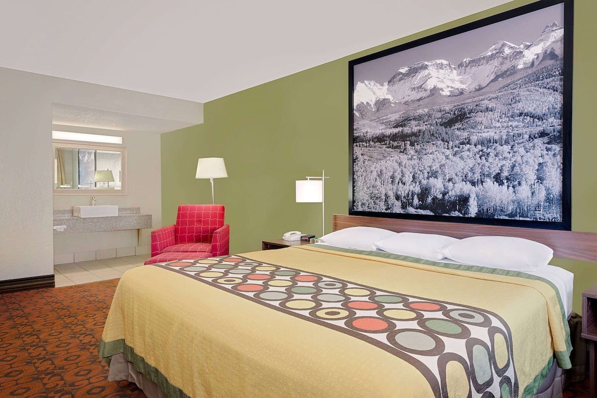 Quarto de hotel com cama, uma poltrona vermelha, uma pintura grande de paisagem montanhosa, banheiro com pia e espelho, e mesa de cabeceira com abajur.
