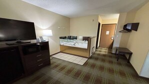 Quarto de hotel com televisão de tela plana, escrivaninha com luminária, banheiro com pia e espelho, e piso coberto por carpete.